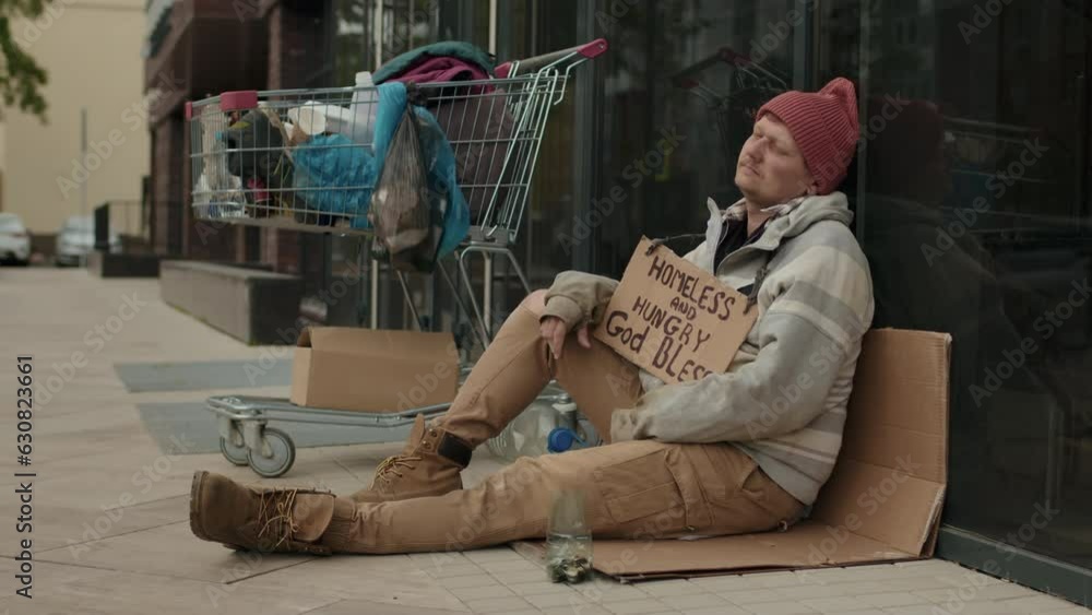 Long shot of tired beggar in rags sitting on cardboard with sign ...