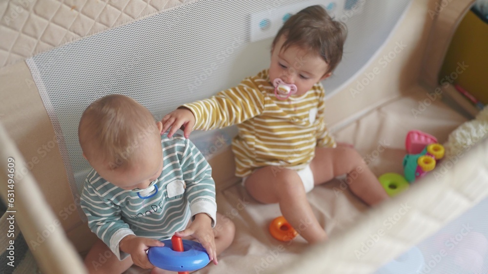 baby twins fight sitting in crib playpen. two baby toddlers are sitting in a crib arena fighting ...