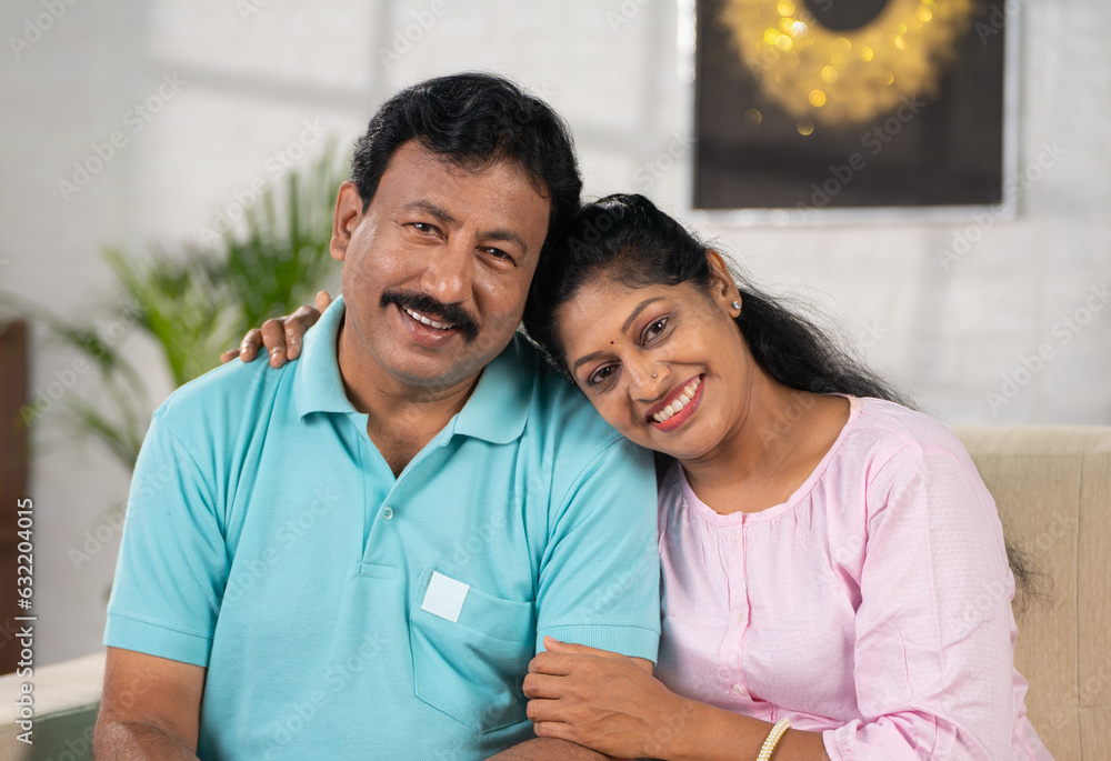 Happy middle aged indian couple looking at camera while sitting on sofa ...
