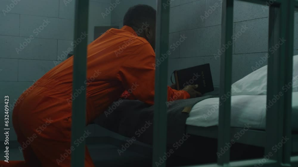 African American prisoner in orange uniform kneels near the bed, prays ...