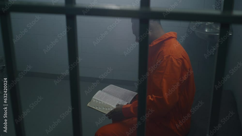African American prisoner in orange uniform sits on bed behind bars ...