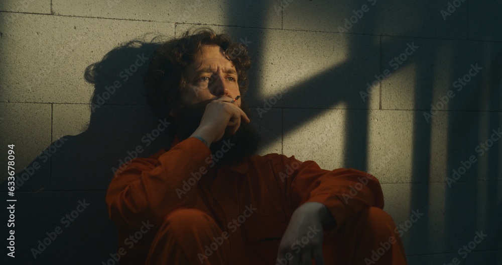 Depressed prisoner in orange uniform sits on bed, smokes cigarette in ...