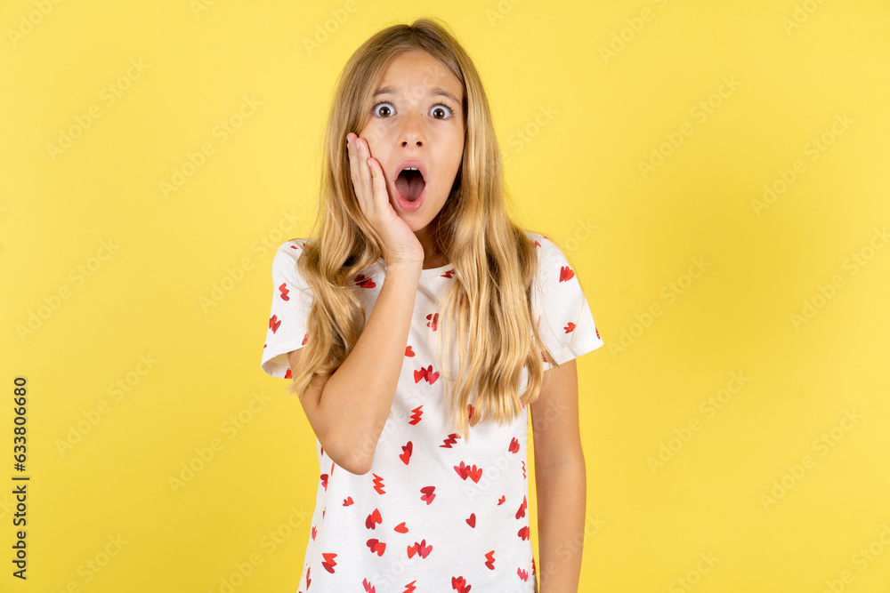 Shocked blonde kid girl wearing polka dot shirt over yellow studio ...