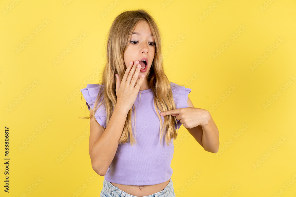 Shocked blonde kid girl wearing violet T-shirt over yellow studio ...