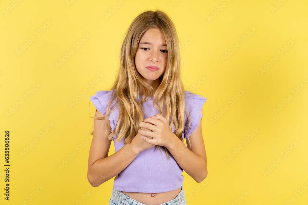 Sad blonde kid girl wearing violet T-shirt over yellow studio ...