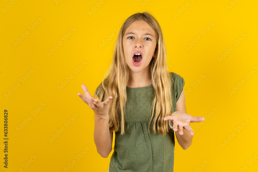 Frustrated blonde kid girl wearing green T-shirt over yellow studio ...