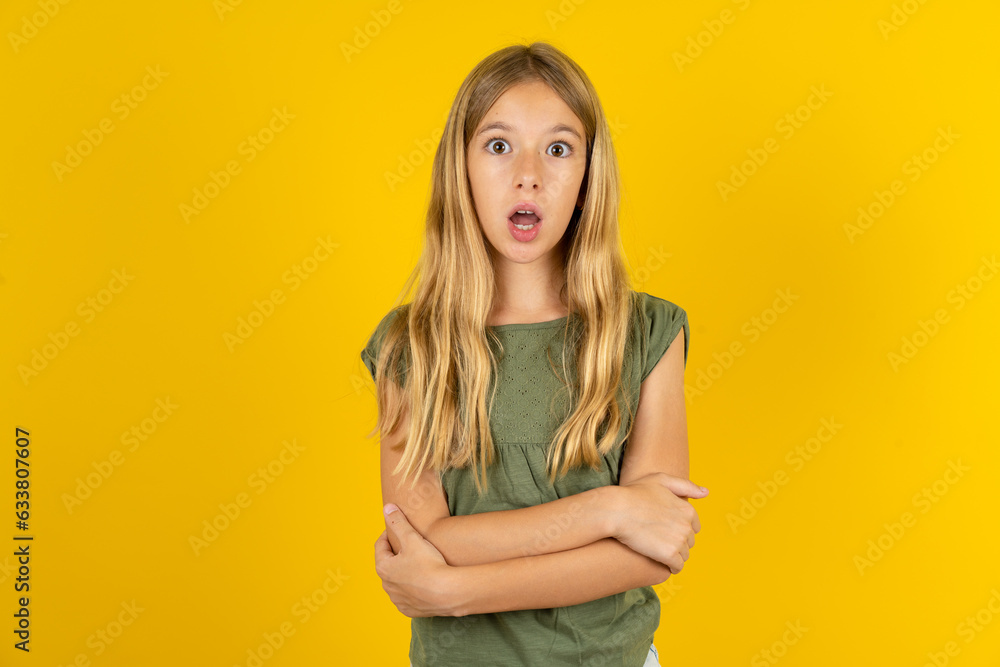 Shocked embarrassed blonde kid girl wearing green T-shirt over yellow ...