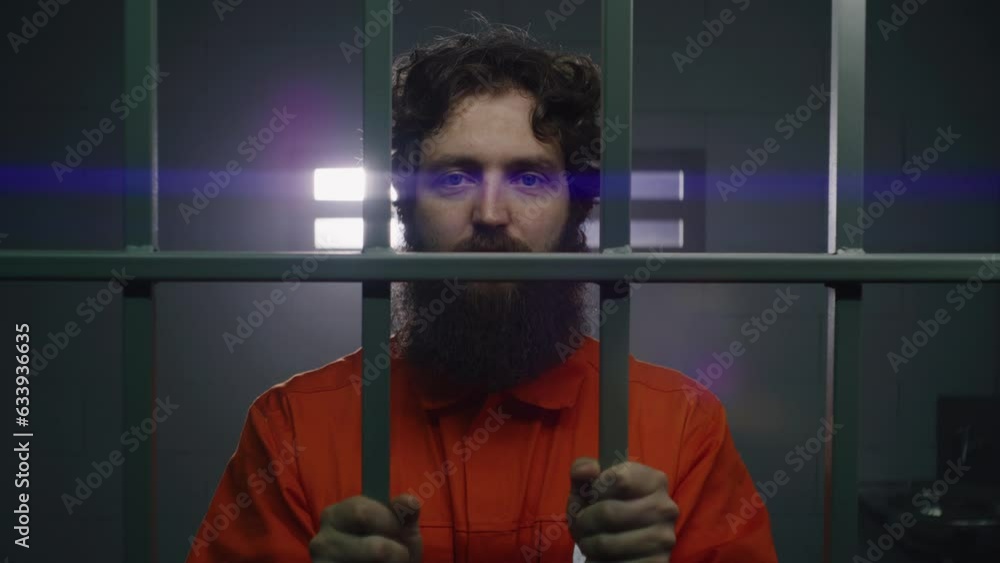 Prisoner in orange uniform holds metal bars, stands in prison cell in ...