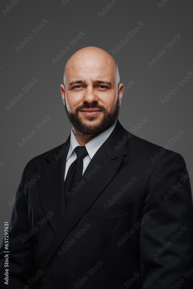 Stately bald man with a full short beard in a black suit and tie poses ...