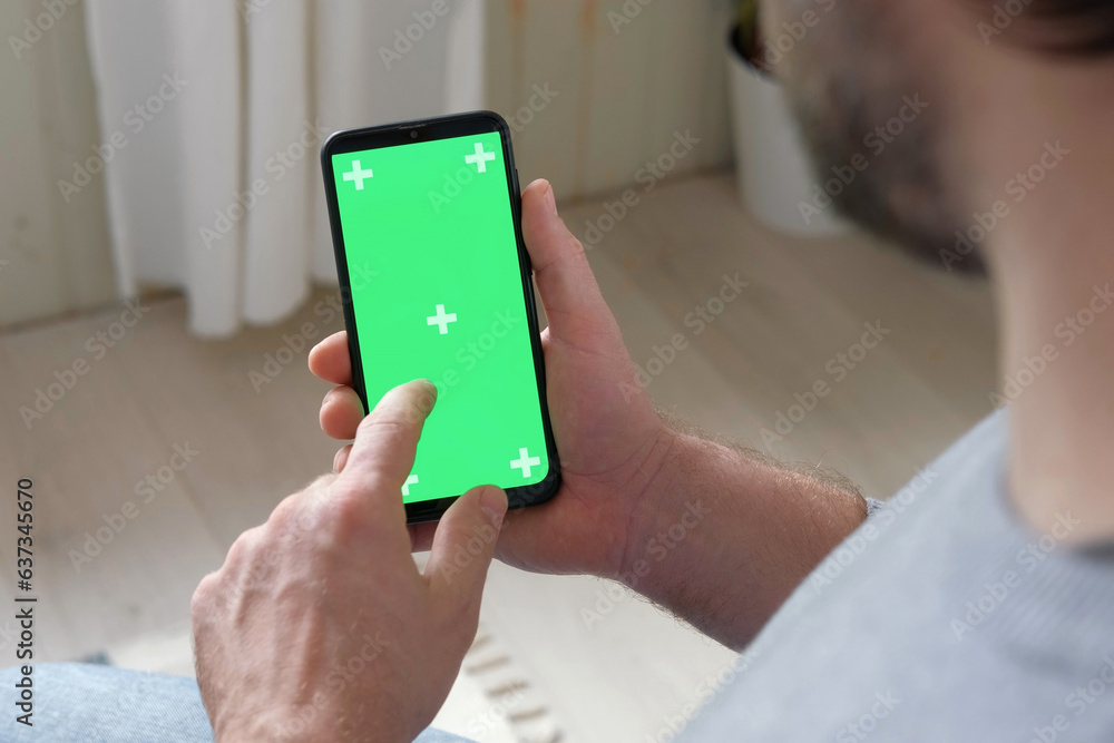 Young man sitting at home holding smartphone green mock-up screen in ...