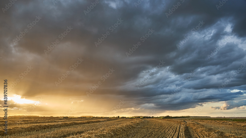 A black rain cloud over a rural field. The rays of the setting sun are ...