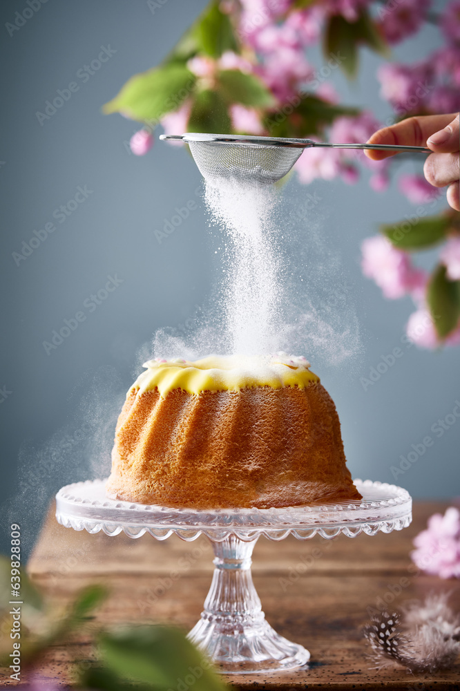 Cropped hand dusting sugar powder on top of the bundt cake. Behind is ...