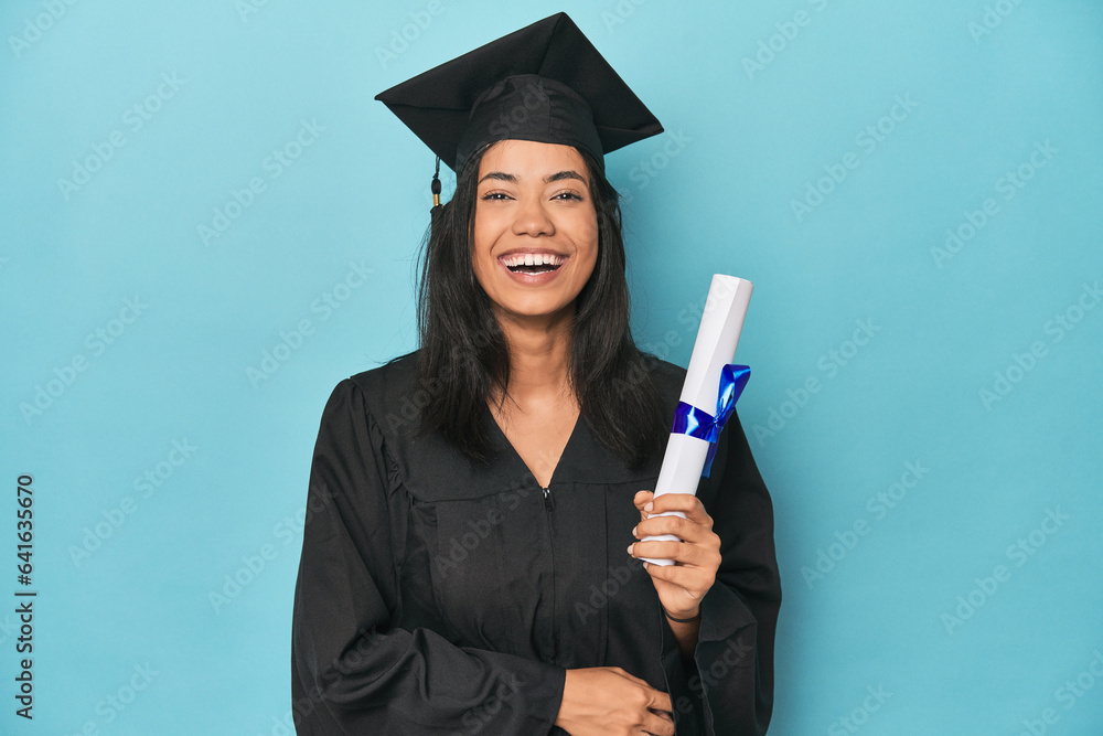 Filipina graduate with diploma on blue studio laughing and having fun ...