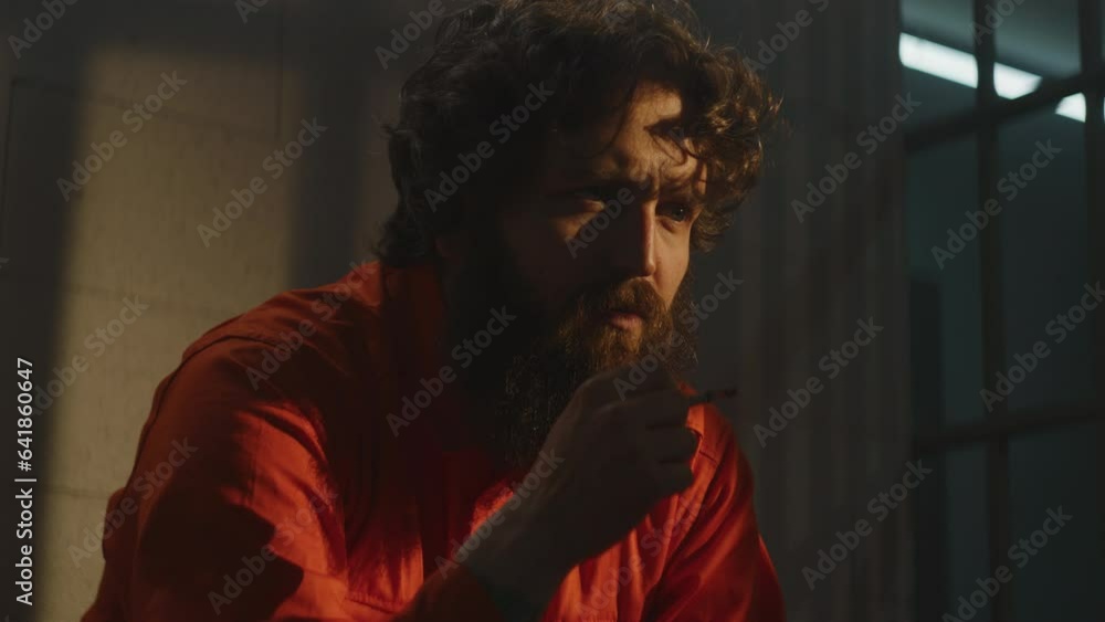 Upset prisoner in orange uniform sits on bed in prison cell, smokes ...