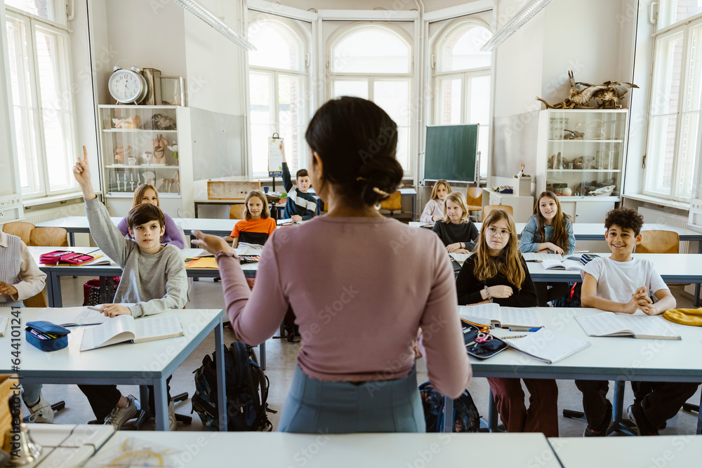 Rear view of female teacher asking students while teaching in classroom ...