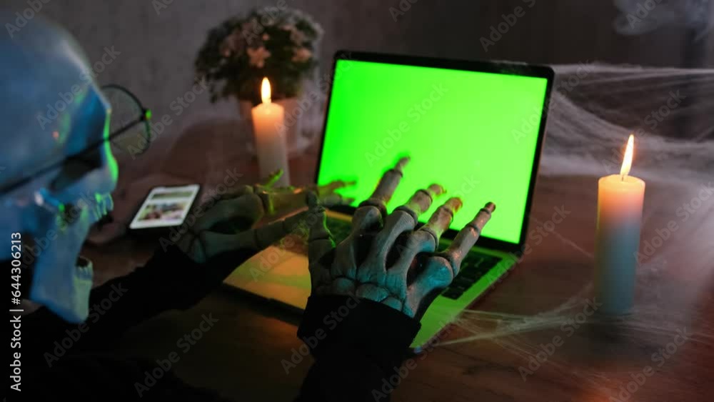 Scary skeleton hands with bony fingers typing keyboard laptop with green screen, surf internet ...
