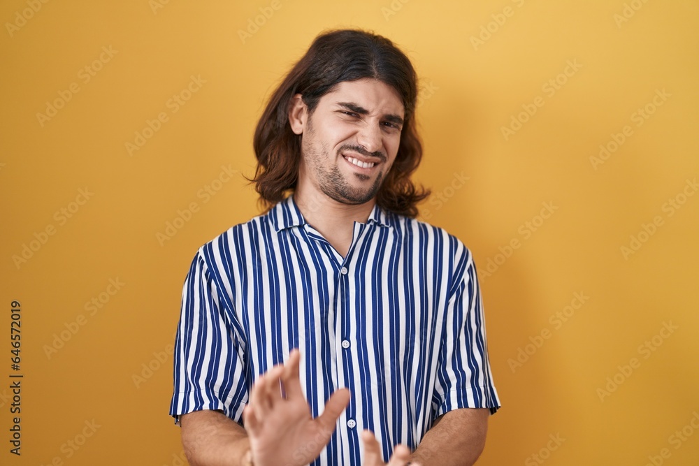 Hispanic man with long hair standing over yellow background disgusted ...