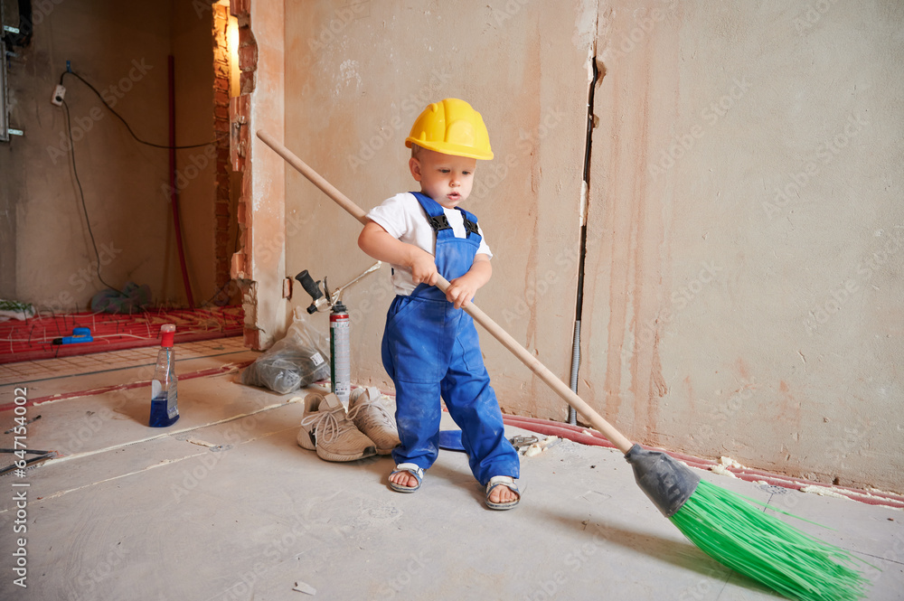 Full length of child in work overalls and safety helmet sweeping floor ...