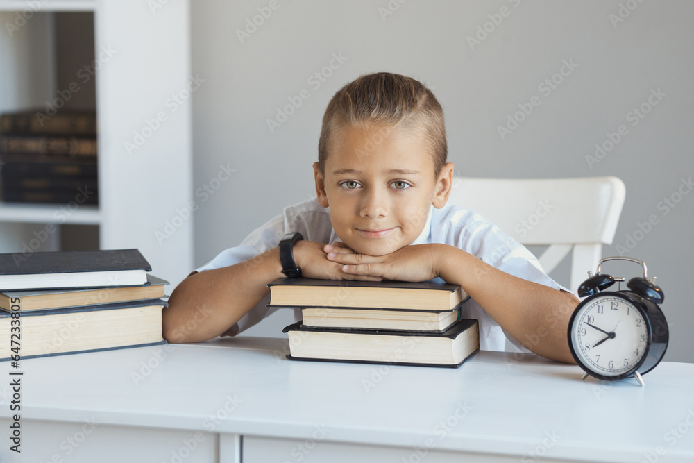 Smart schoolboy in a white shirt sits at a desk with a large stack of ...