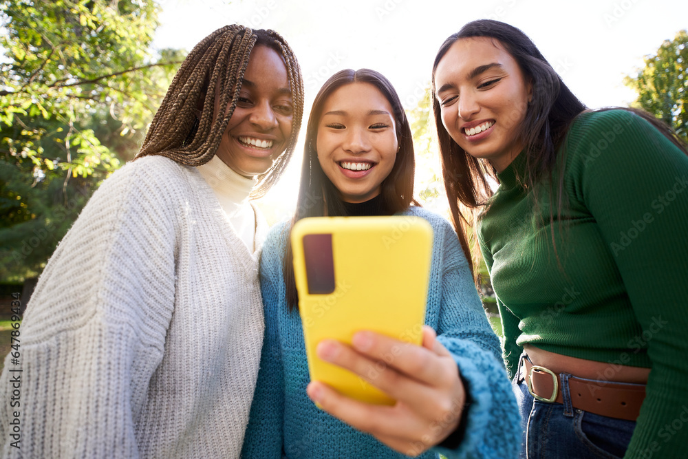 Three smiling multiracial young girls using cell phone in outdoor park ...