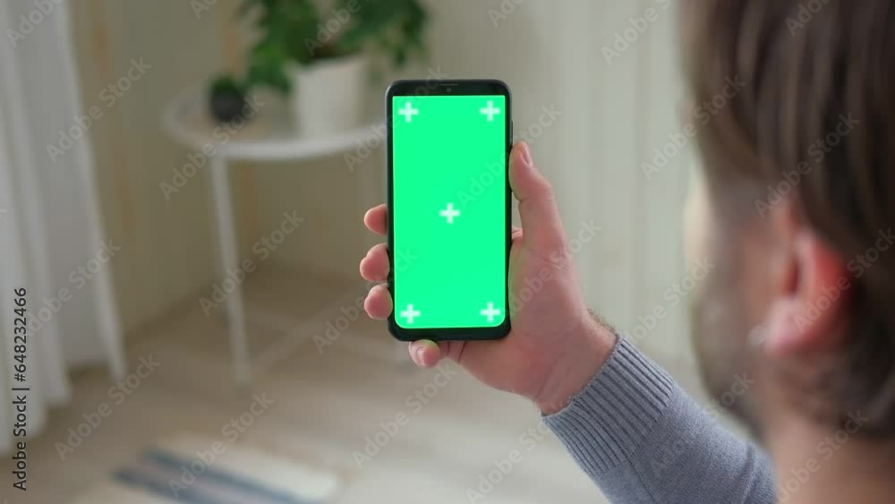 Young man sitting at home holding smartphone greenscreen mock-up in hand. Male person using ...