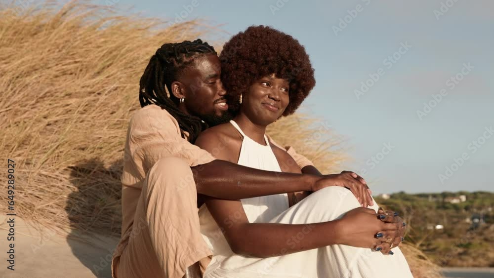 A stylish black African couple, in a romantic moment, sits on a sandy ...