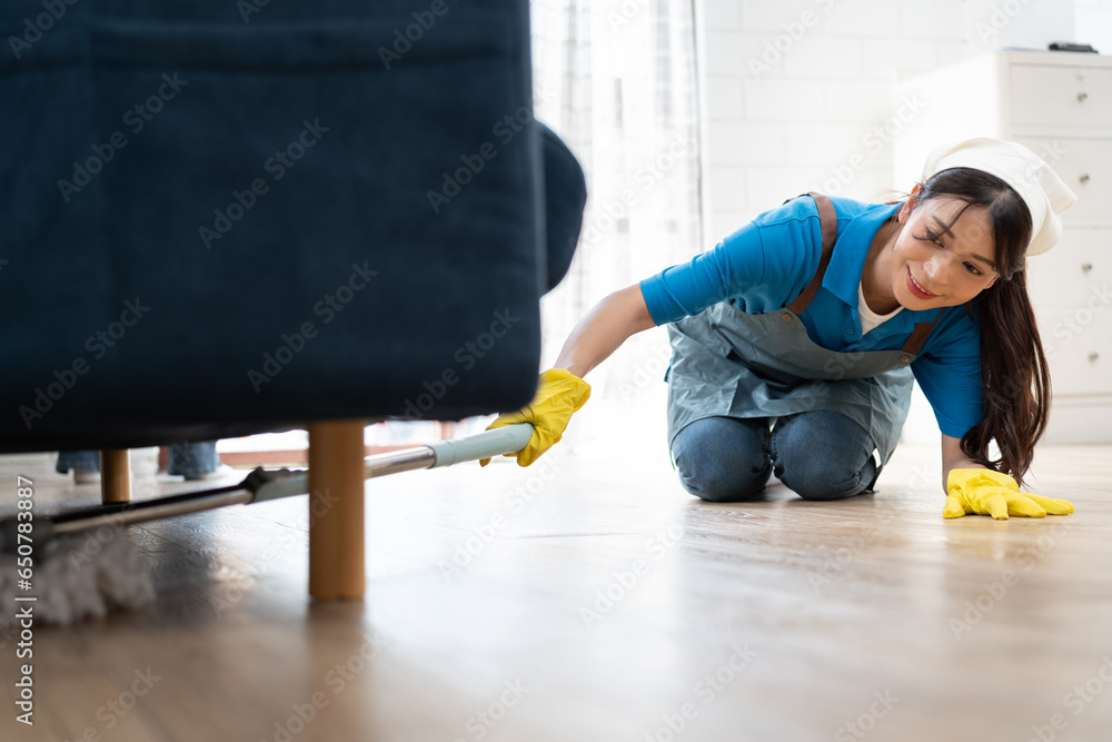 Asia young woman in maid uniform cleaning under sofa floor with wet mop ...
