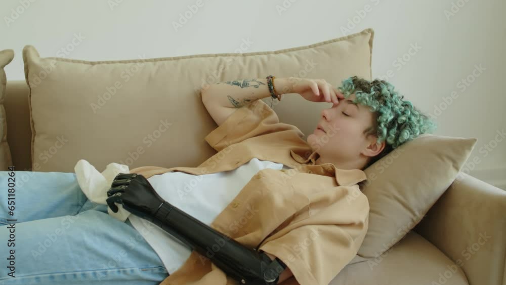 Sad gen Z girl with prosthetic arm and blue hair lying on sofa, holding ...