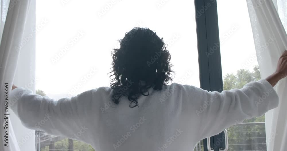 Biracial woman wearing bathrobe opening curtains and looking through ...