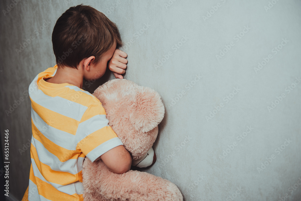 A little boy cries in the gray corner, hiding his face and hugs the ...