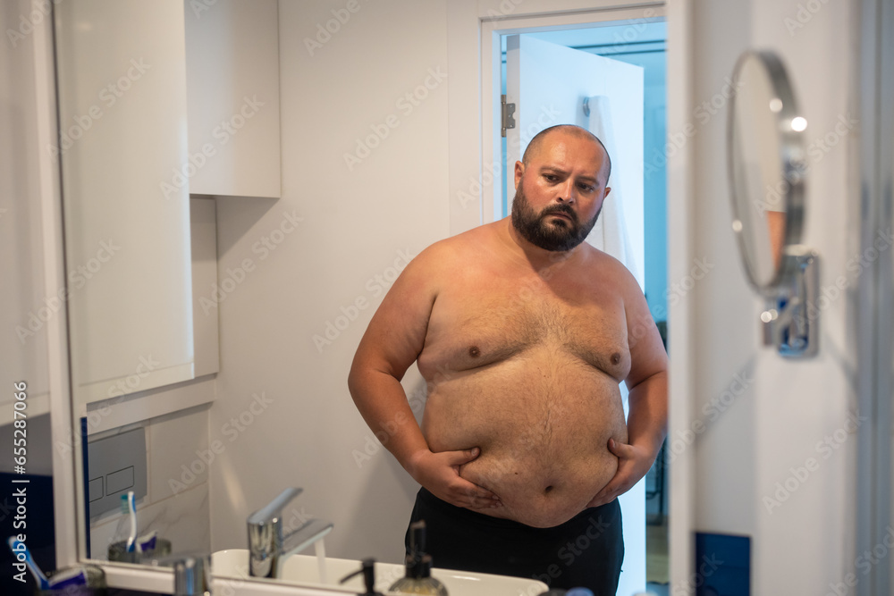 Fat obese guy touching stomach in front of mirror. Young bearded bald ...