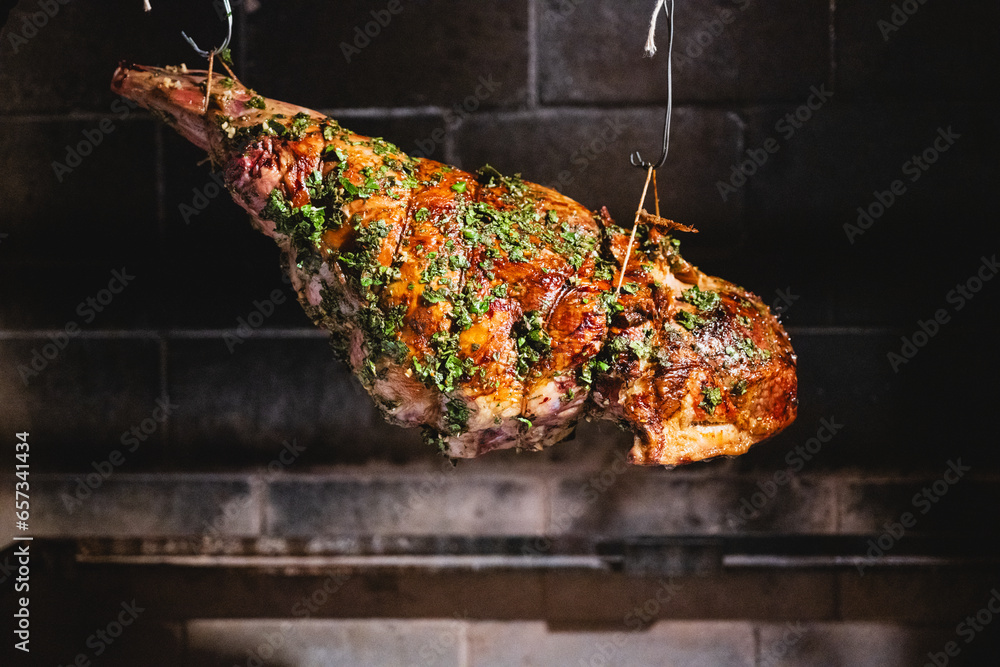 A leg of lamb covered in herbs hangs roasting over hot coals in a ...
