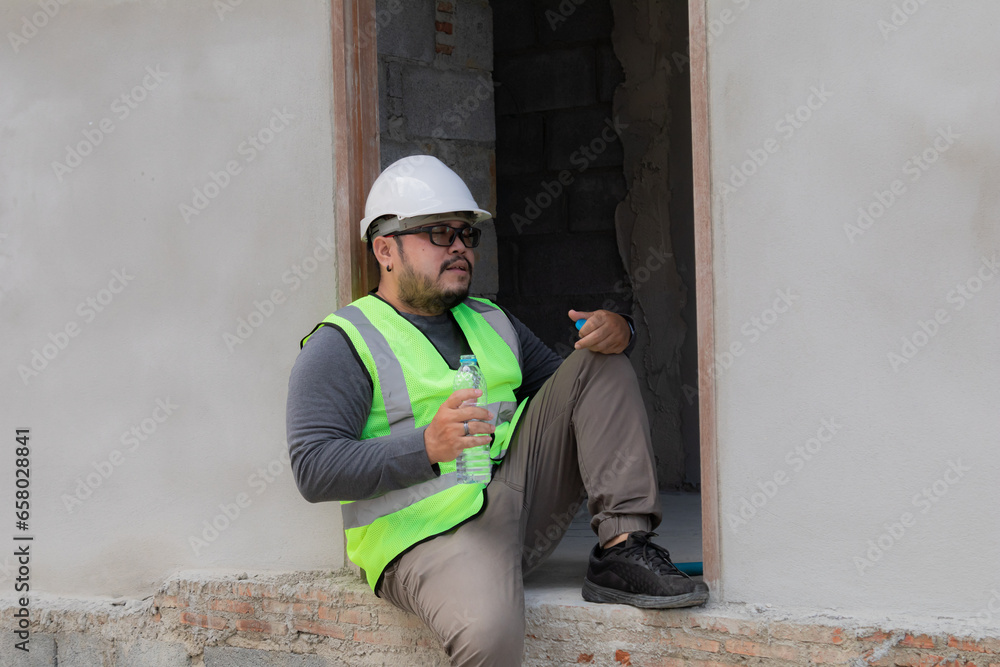 Young Asian engineering worker overweight employee foreman tired sitting on construction site ...
