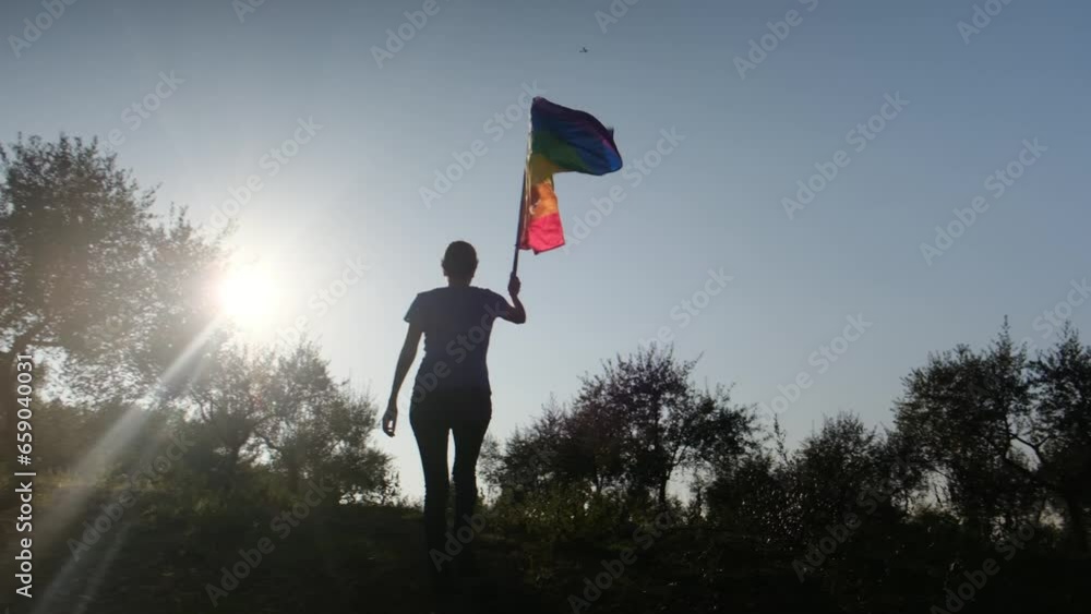 Young woman alone with LGBTQ+ flag walking as it waves in the sunset ...