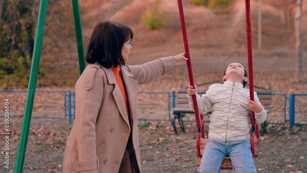 Happy family. Little son sitting on swing smiling, mom rocking child on ...