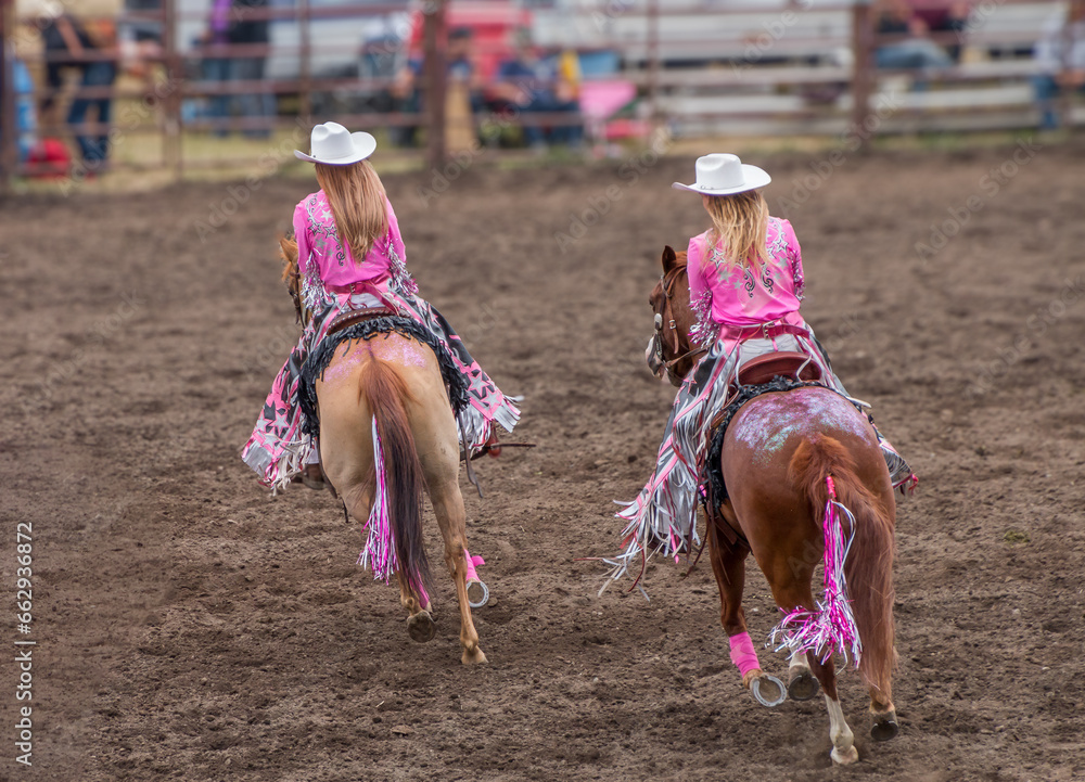 2 cowgirls riding two horse at a rodeo in a dirt arena. The horses are ...