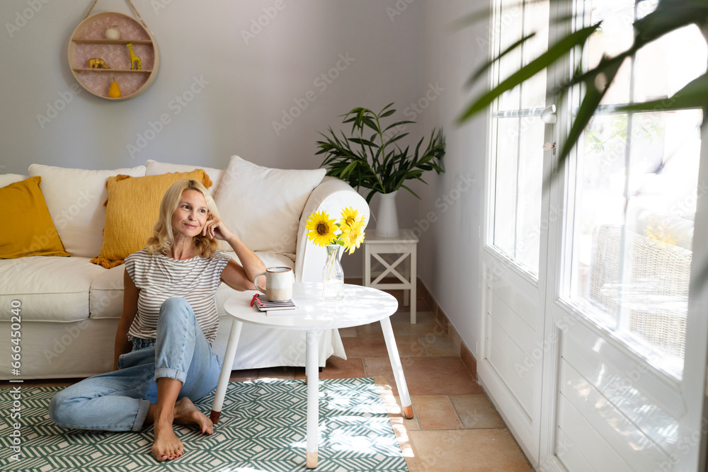 Contemplative woman sitting on floor by table in living room at home ...