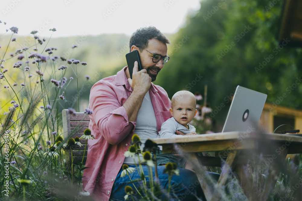Father holding baby while working on laptop outdoors, in garden ...