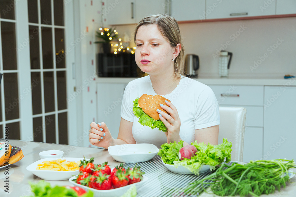 Young woman eating hamburger in the kitchen with full table of different food, problems with ...