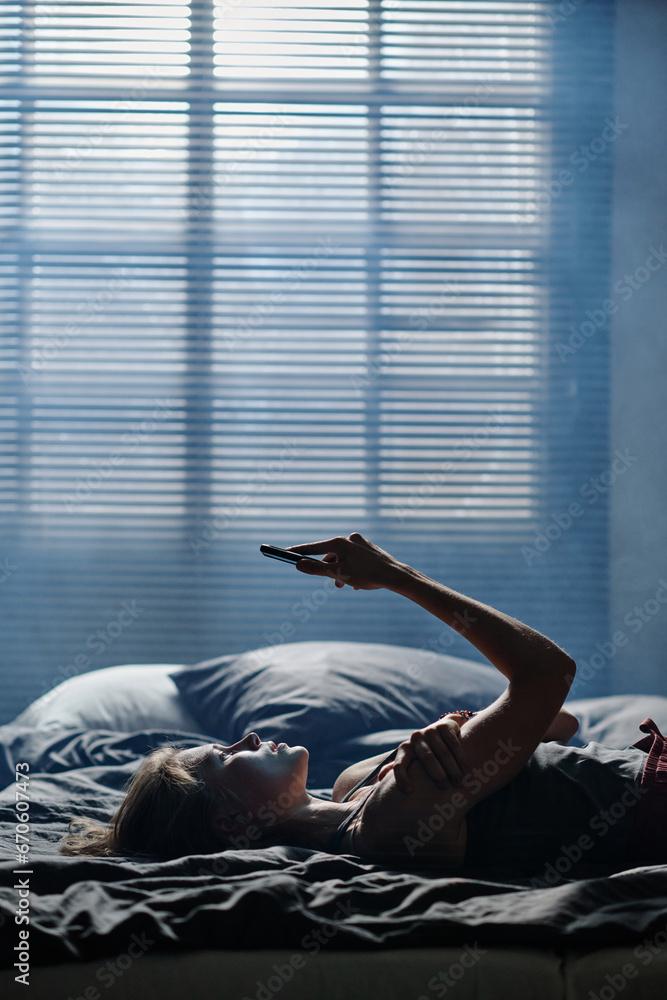 Offended teenage girl with mobile phone lying on bed against window ...