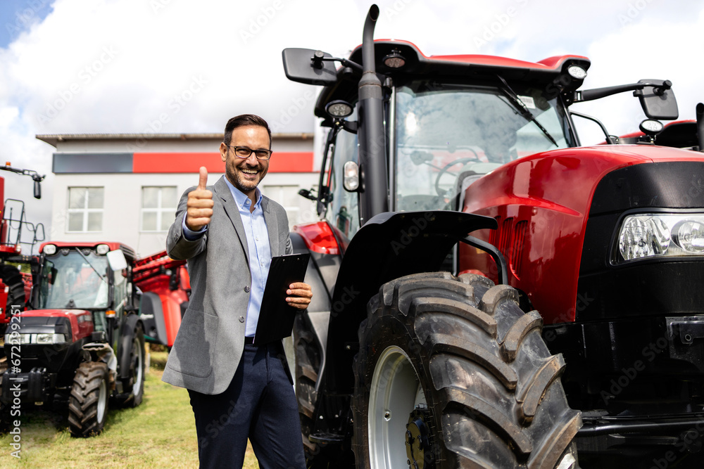 Portrait of successful tractor dealer standing by farming equipment and ...