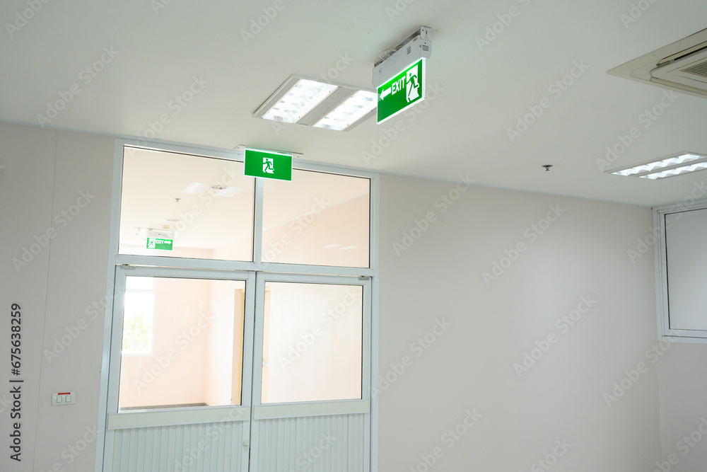 Selective fire exit sign on white ceiling and glass door.Green fire ...