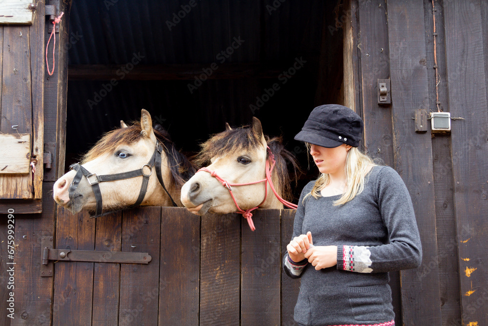 Pretty teenage girl stands by wooden stable door as two look a like dun ...