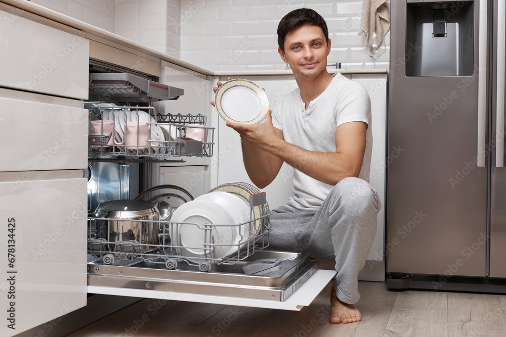 handsome positive man takes out clean dishes from the dishwasher in ...