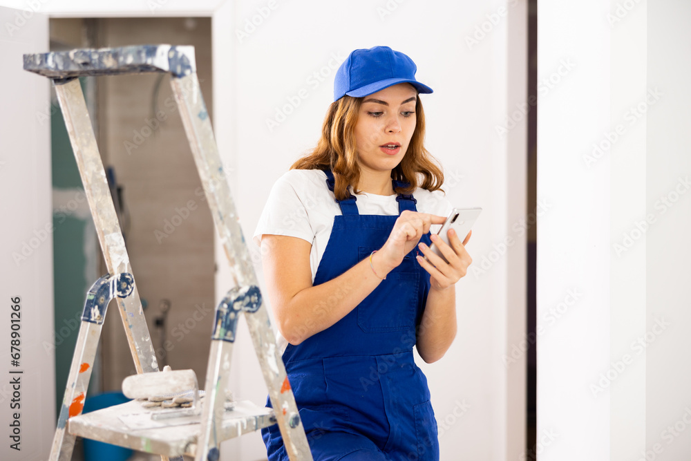 Puzzled surprised young female builder in blue overall and cap reading ...