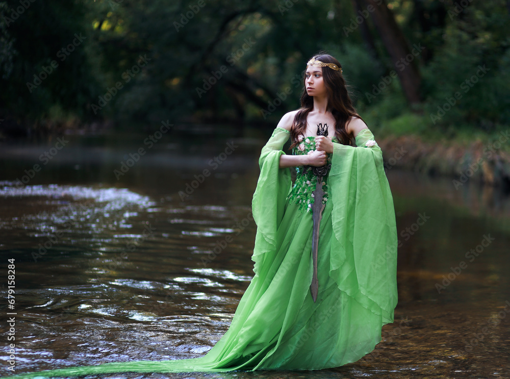 Young fairy woman in long green dress standing in river with sword in ...