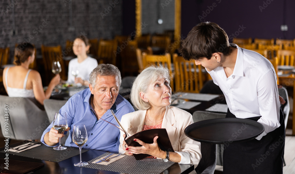 Polite hospitable young waiter talking to positive elderly couple, man ...