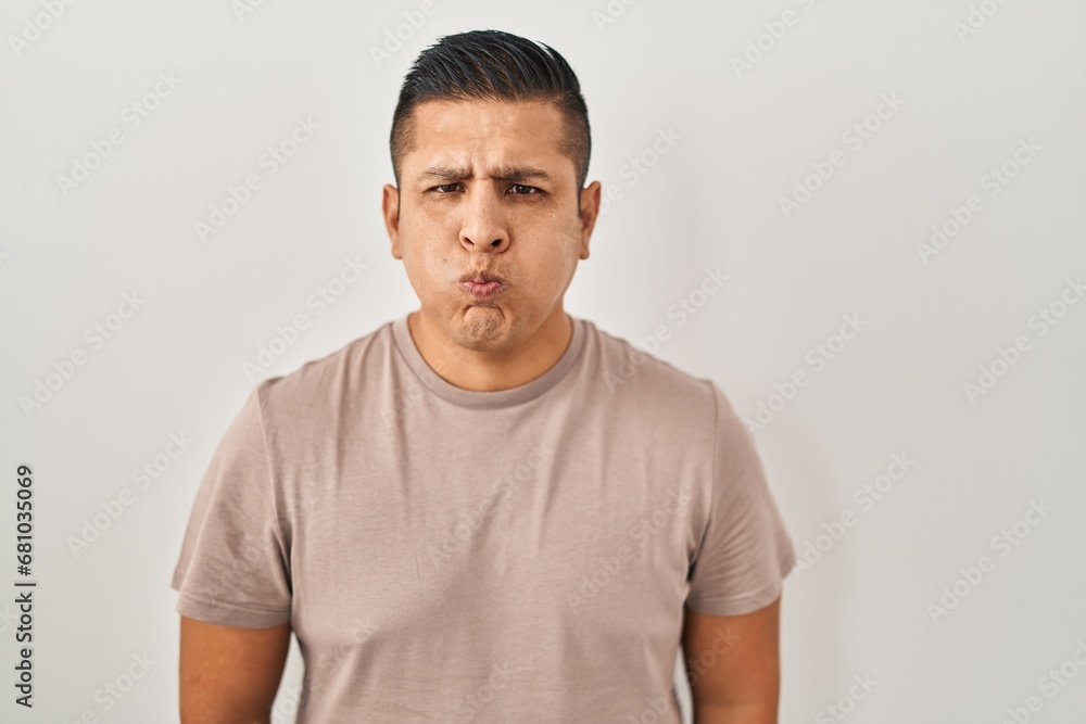 Hispanic young man standing over white background puffing cheeks with ...