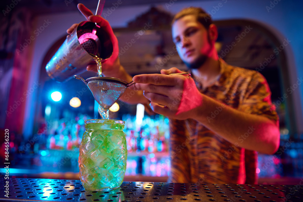 Attractive young bartender gently pouring refreshing mojito from shaker into cocktail glass on ...