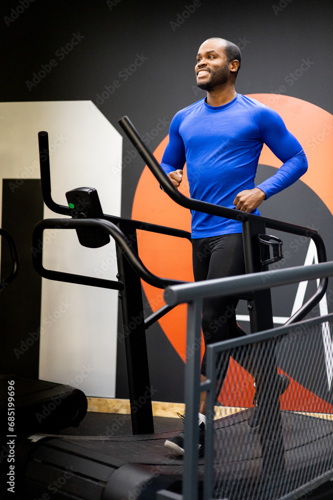 An African man in sportswear runs on a curved treadmill inside a gym ...